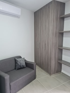 Um quarto com sofá cinza, almofada tecida cinza, guarda-roupa cinza com espelho e estante cinza.