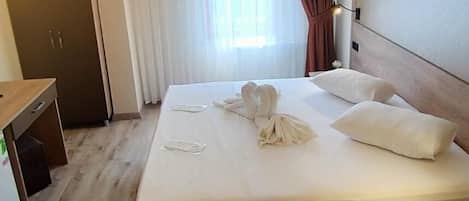 Uma cama bem arrumada com lençóis e almofadas brancas, uma escrivaninha de madeira com uma cadeira e uma janela com cortinas leves.