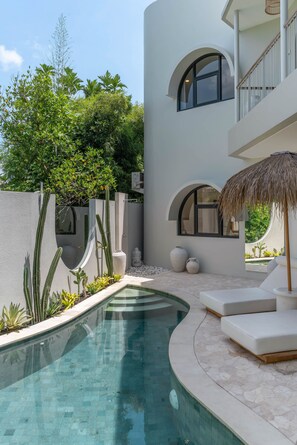 Uma área de piscina moderna com design curvo, cercada por paredes brancas e um guarda-sol de palha.