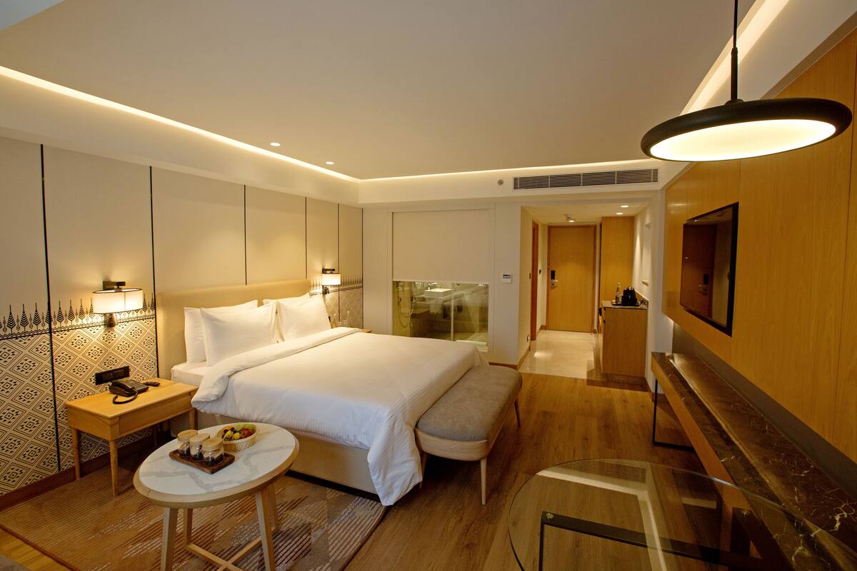 Quarto de hotel moderno com uma cama grande, mesas de cabeceira, uma pequena mesa redonda com uma bandeja de petiscos, um banco, uma mesa de centro de vidro e uma TV fixada na parede.