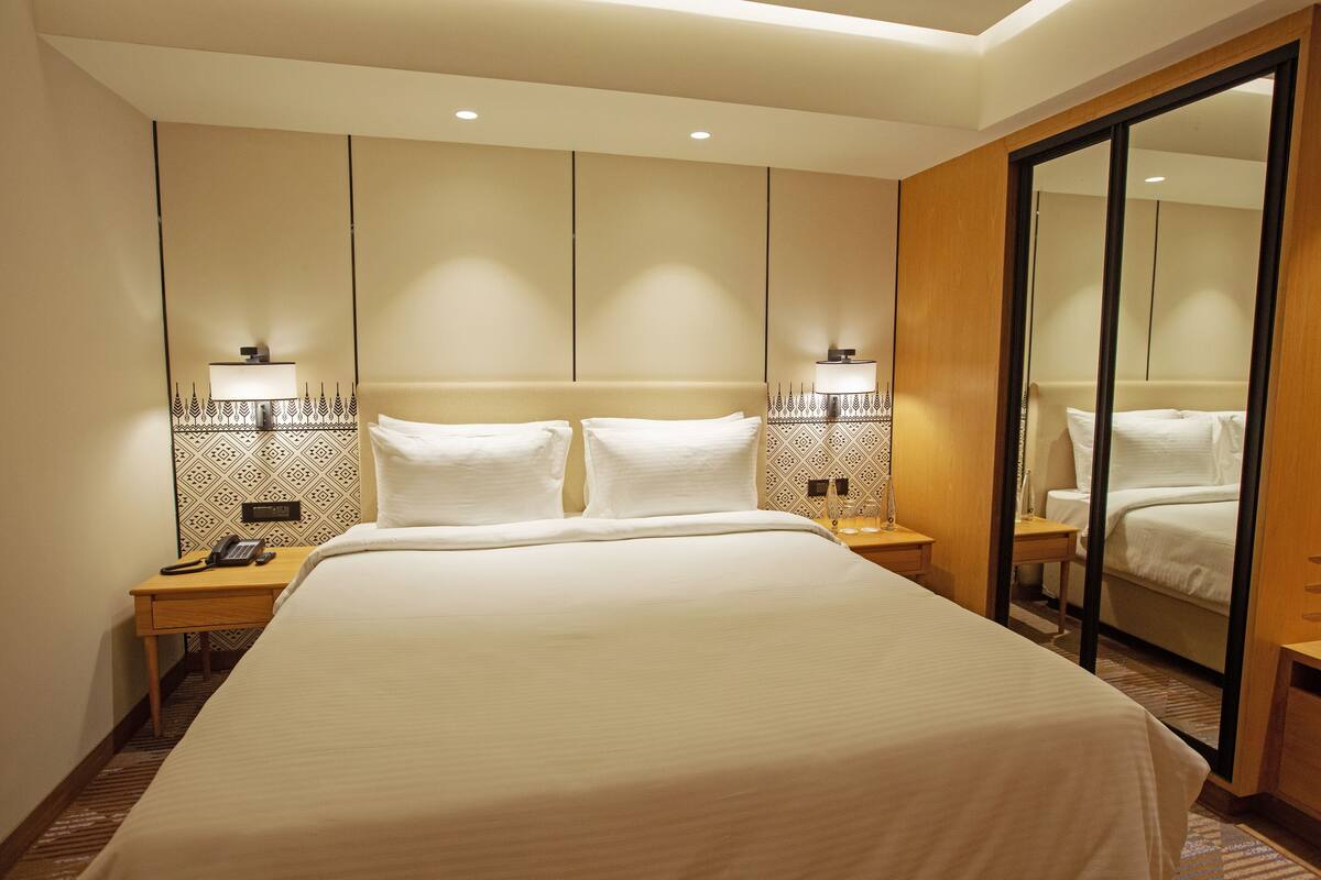 Quarto de hotel com uma cama grande, duas mesas de cabeceira com abajures, um espelho e um guarda-roupa de madeira.