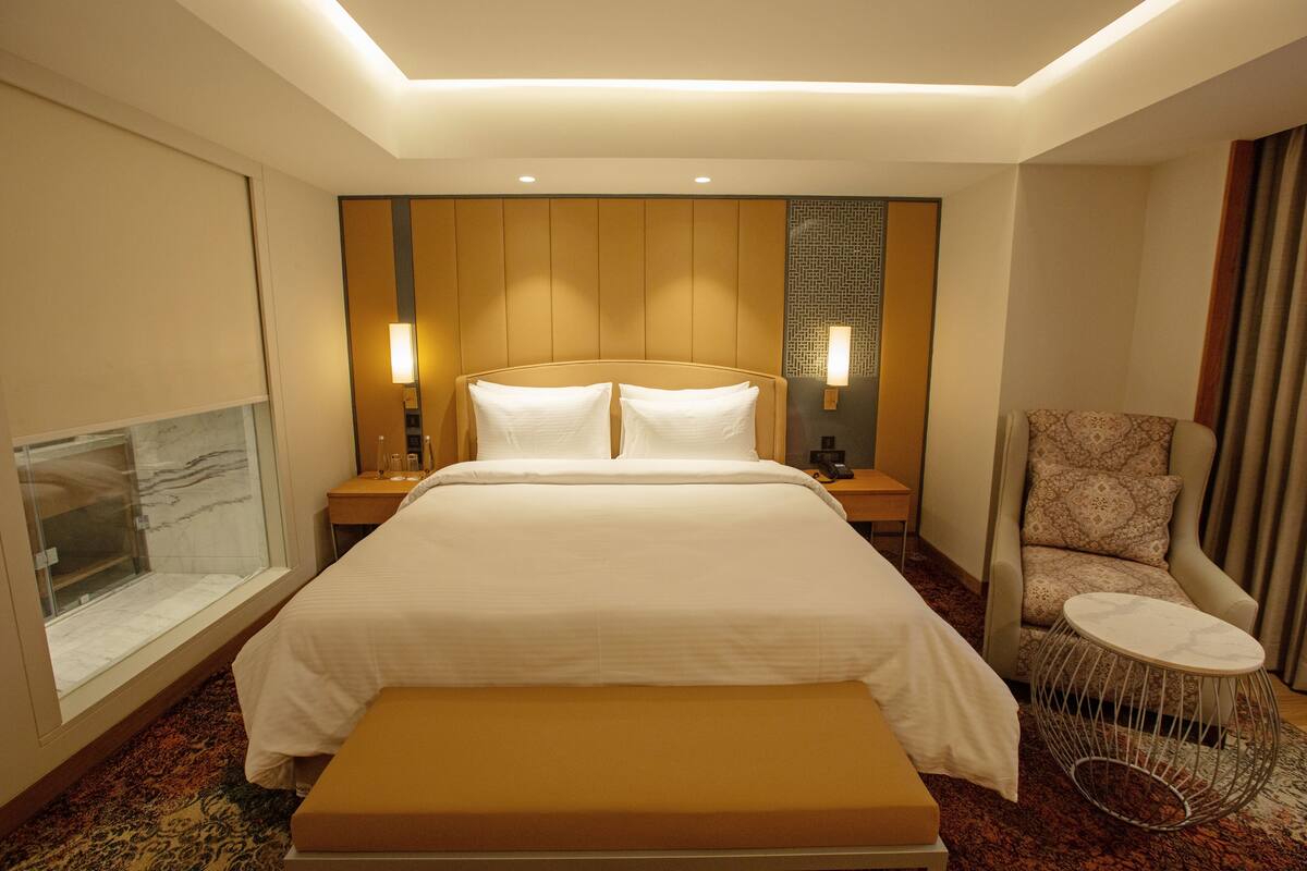 Quarto de hotel com uma cama grande, mesas de cabeceira, uma cadeira, uma pequena mesa redonda e uma janela com proteção de vidro.