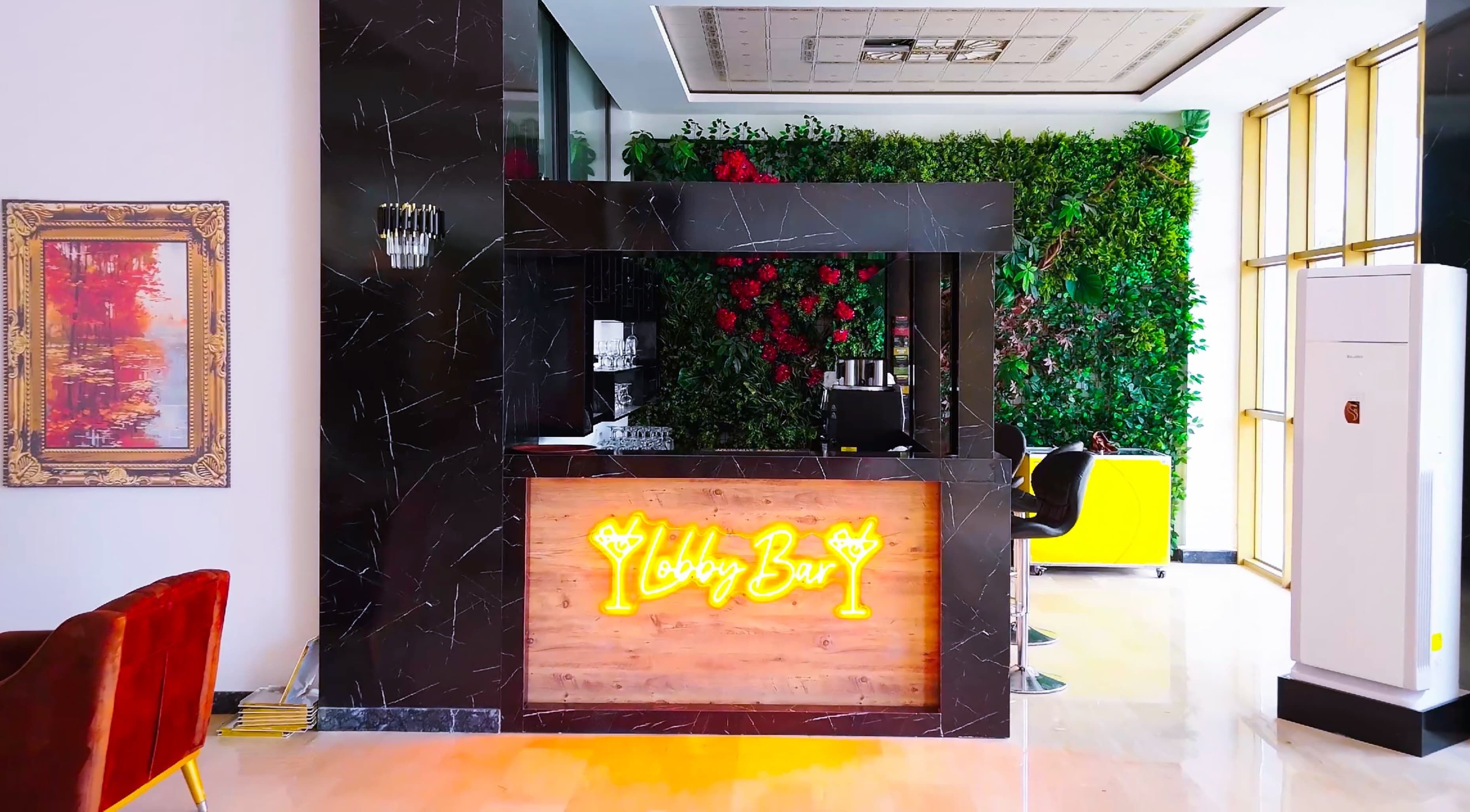 Um lobby com balcão de recepção, uma placa de neon que diz "Lobby Bar", um quadro emoldurado e uma parede verde com plantas.