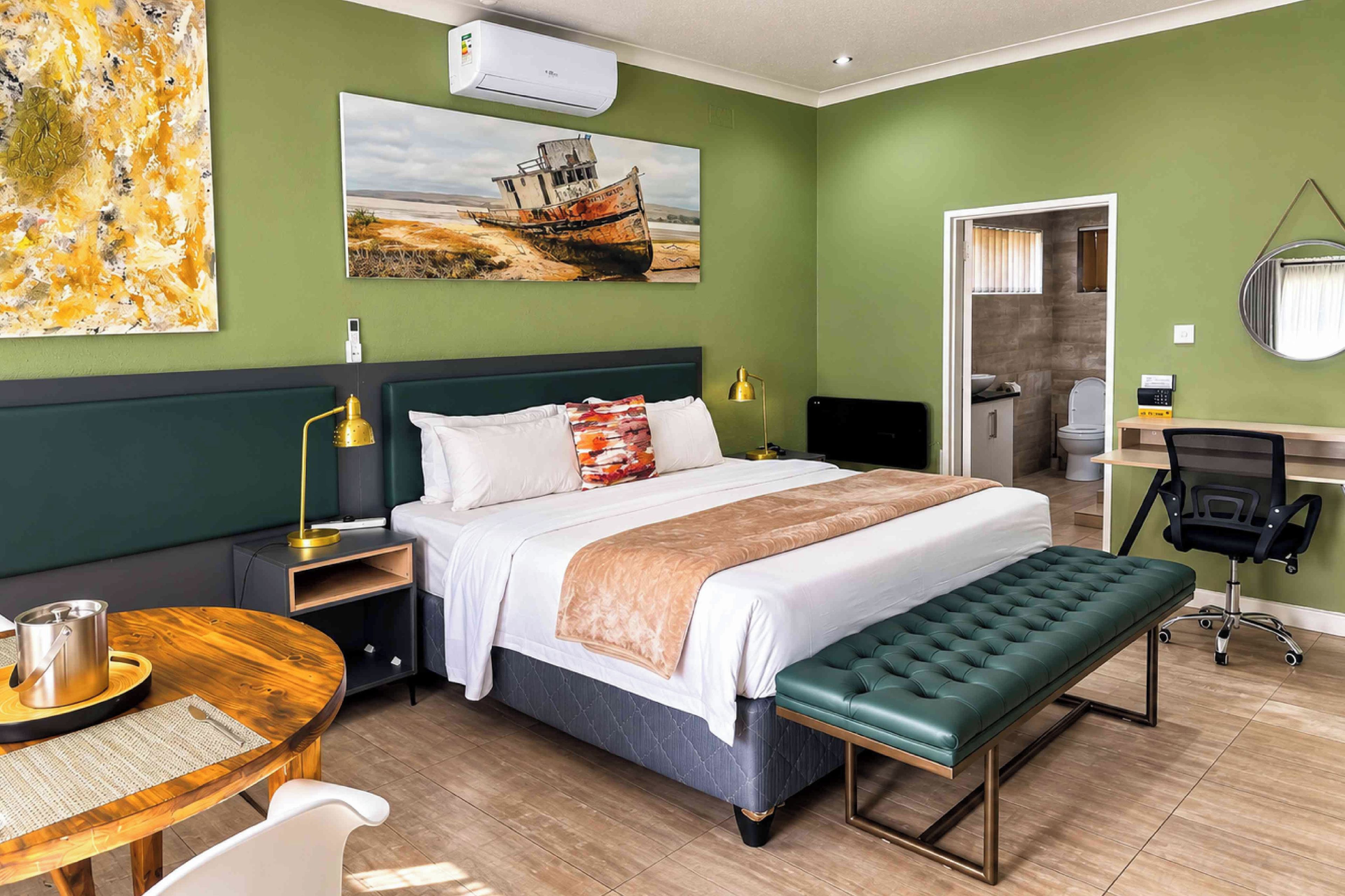 Quarto de hotel com cama, mesa de cabeceira, luminária, banco, escrivaninha, cadeira e um quadro na parede.