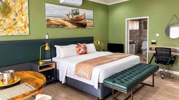 Quarto de hotel com cama, mesa de cabeceira, luminária, banco, escrivaninha, cadeira e um quadro na parede.