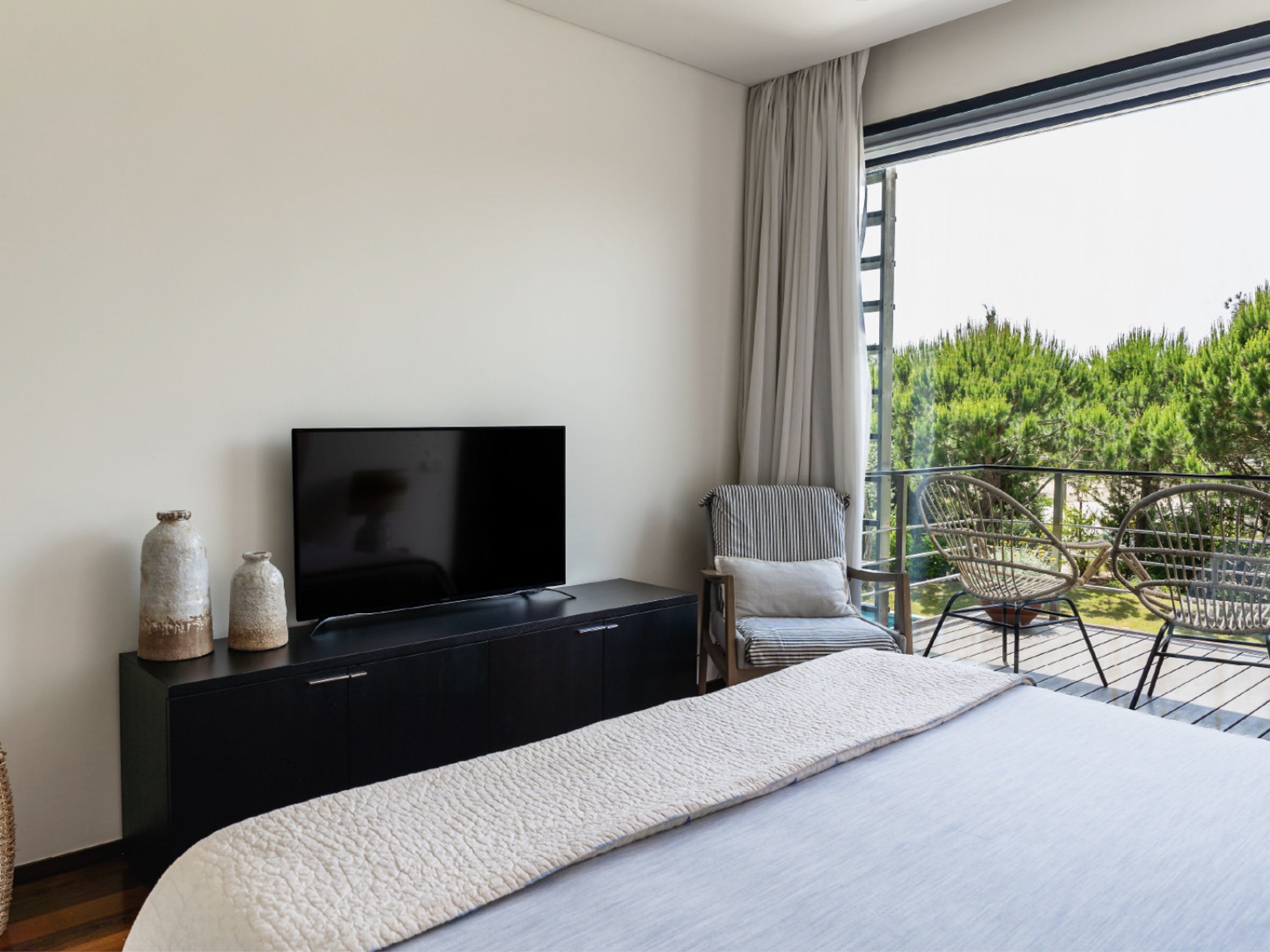 Quarto moderno com televisão de tela plana sobre uma cômoda preta, uma cama grande com manta branca texturizada, uma cadeira de vime e uma varanda com vista para árvores.