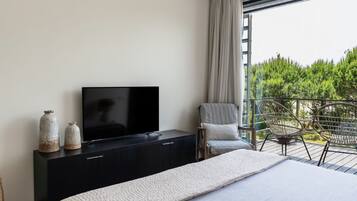 Quarto moderno com televisão de tela plana sobre uma cômoda preta, uma cama grande com manta branca texturizada, uma cadeira de vime e uma varanda com vista para árvores.