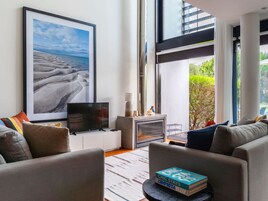 Uma sala de estar moderna com uma TV grande, assentos confortáveis, uma estante de livros e vista para o verde através de amplas janelas.