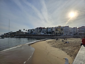 Uma praia costeira com um muro de pedra, um veleiro e edifícios ao fundo.