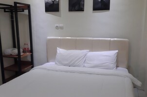 Cama bem arrumada com lençóis brancos em quarto de hotel. Há uma prateleira de madeira com toalhas e artigos de higiene pessoal.