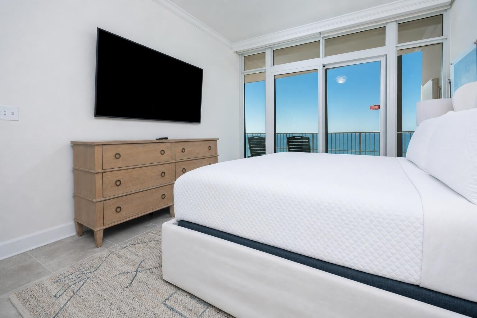 Quarto com cama, penteadeira, televisão e janelas amplas com vista.