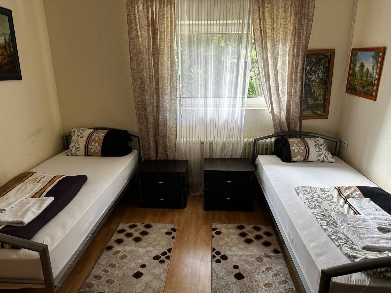 Um quarto pequeno e bem iluminado, com duas camas de solteiro, uma janela com cortinas finas e vista para árvores.