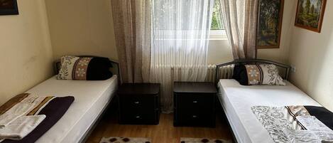 Um quarto pequeno e bem iluminado, com duas camas de solteiro, uma janela com cortinas finas e vista para árvores.