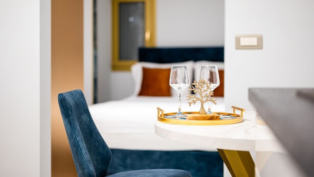 Quarto de hotel com uma mesa de jantar posta com dois copos e um tabuleiro, uma cama com cabeceira e um quadro na parede.