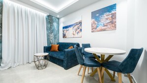 Uma sala de estar moderna com um sofá azul, uma mesa redonda e duas cadeiras. Há duas pinturas na parede e uma pequena mesa redonda com um design geométrico.