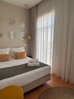 Quarto de hotel moderno com uma cama grande, uma mesa redonda pequena e vista da cidade por cortinas translúcidas.