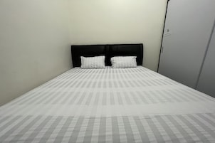 Uma cama de solteiro com cabeceira preta e duas almofadas, parede lisa e uma porta à direita.