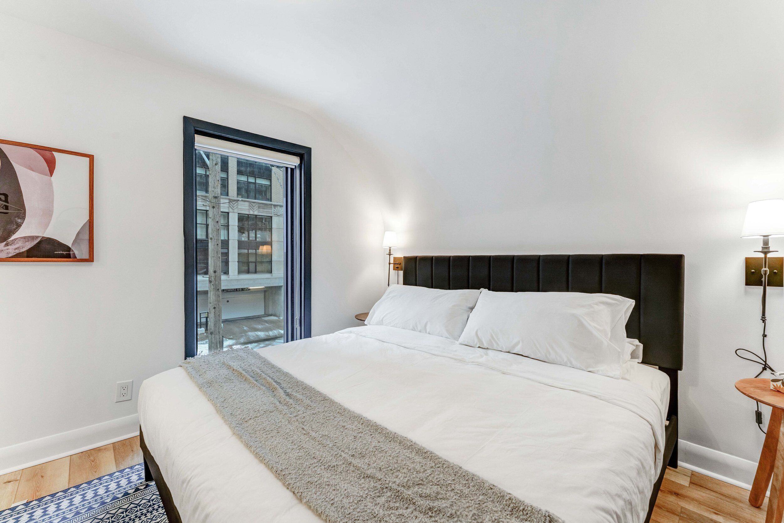 Quarto de hotel moderno com uma cama grande, vista através de uma porta de vidro e uma pequena mesa de cabeceira com abajur.