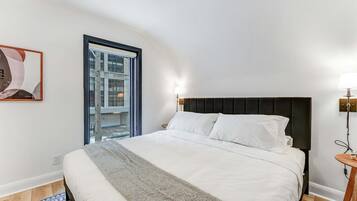 Quarto de hotel moderno com uma cama grande, vista através de uma porta de vidro e uma pequena mesa de cabeceira com abajur.