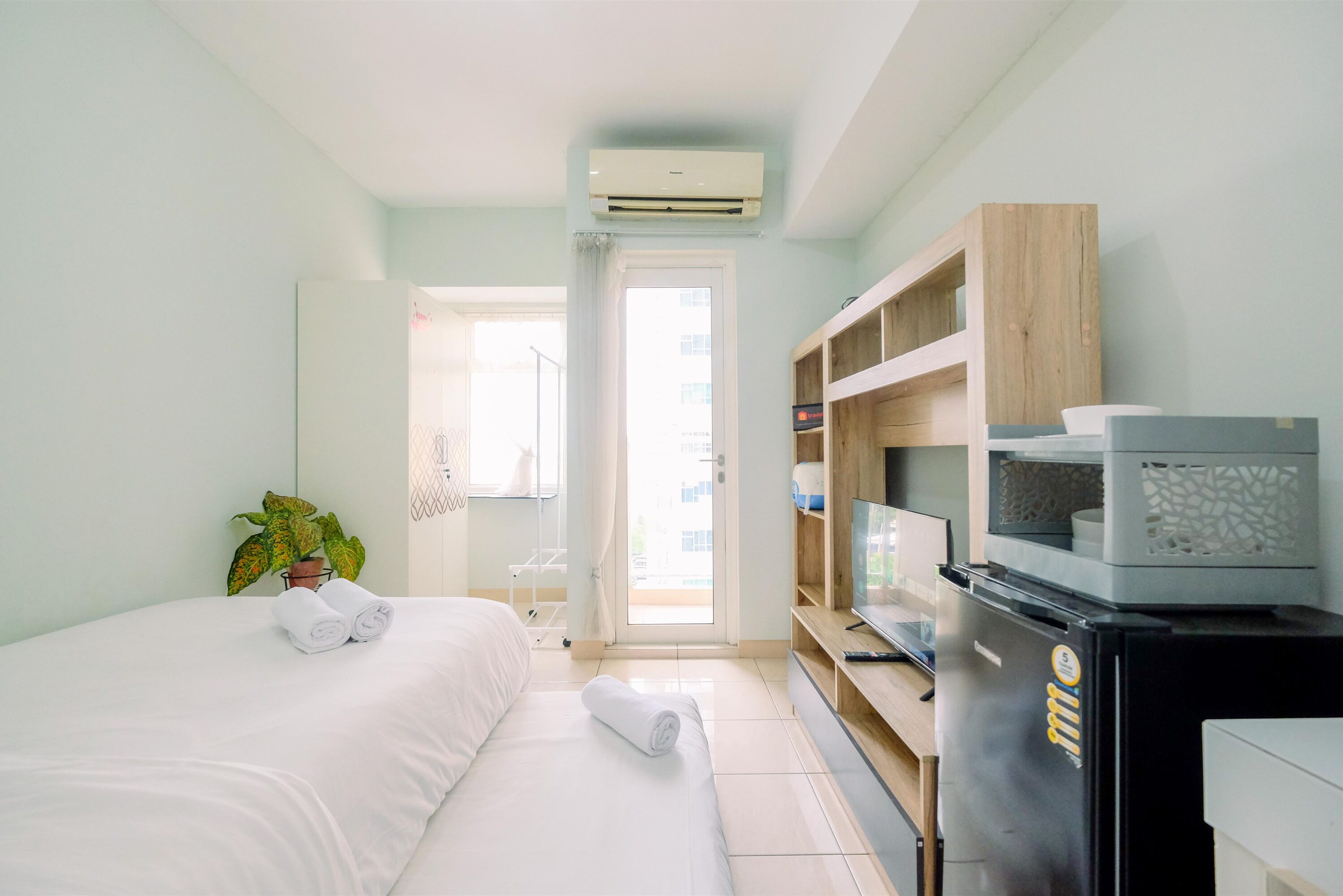 Quarto de hotel com cama, escrivaninha, frigorífico e uma planta.