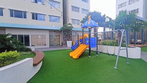 Um parquinho com escorregador e balanços, cercado por grama sintética e plantas, em frente a um edifício com grandes janelas e varandas.
