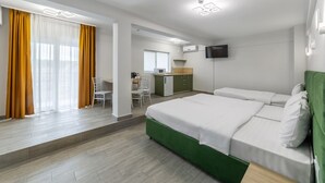 Quarto de hotel com cama verde, cortinas brancas, secretária com cadeiras, cozinha compacta e televisão na parede.