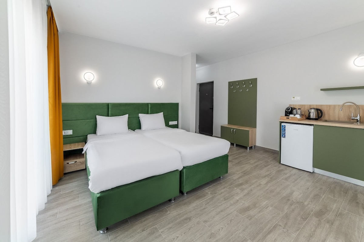 Quarto de hotel moderno com cama verde, roupa de cama branca, uma kitchenette pequena e banheiro separado.