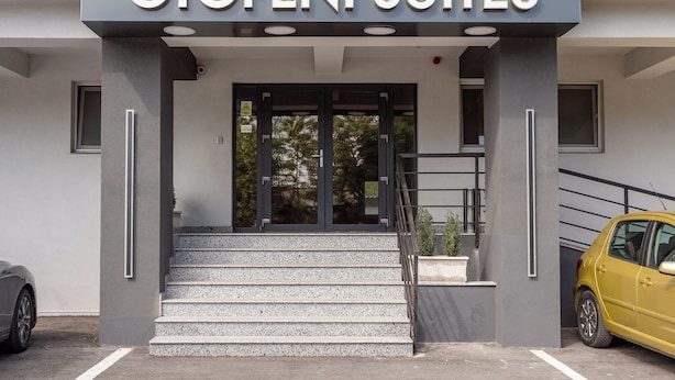 Entrada do Otopeni Suites, com um conjunto de degraus que levam à porta, um carro amarelo estacionado à direita e fachada de um edifício moderno.