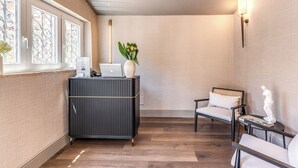 Uma área de recepção com um console preto, um vaso branco com uma planta, uma cadeira de vime e uma estátua sobre uma mesinha.