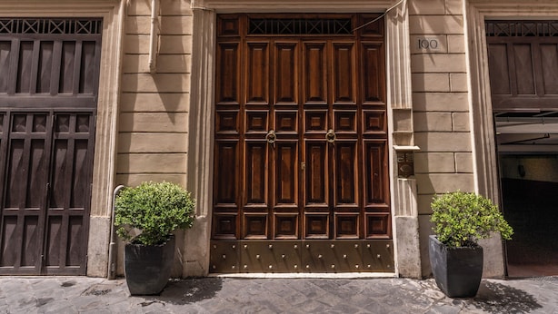Entrada de um edifício com uma grande porta de madeira, duas plantas em vasos e uma placa com um número acima da porta.