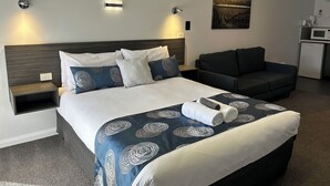 Quarto de hotel com uma cama grande, cabeceira escura, mesa de cabeceira pequena, sofá escuro, micro-ondas e frigorífico.