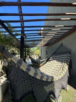 Uma rede sob uma pérgola de madeira, cercada por palmeiras e um muro de concreto.