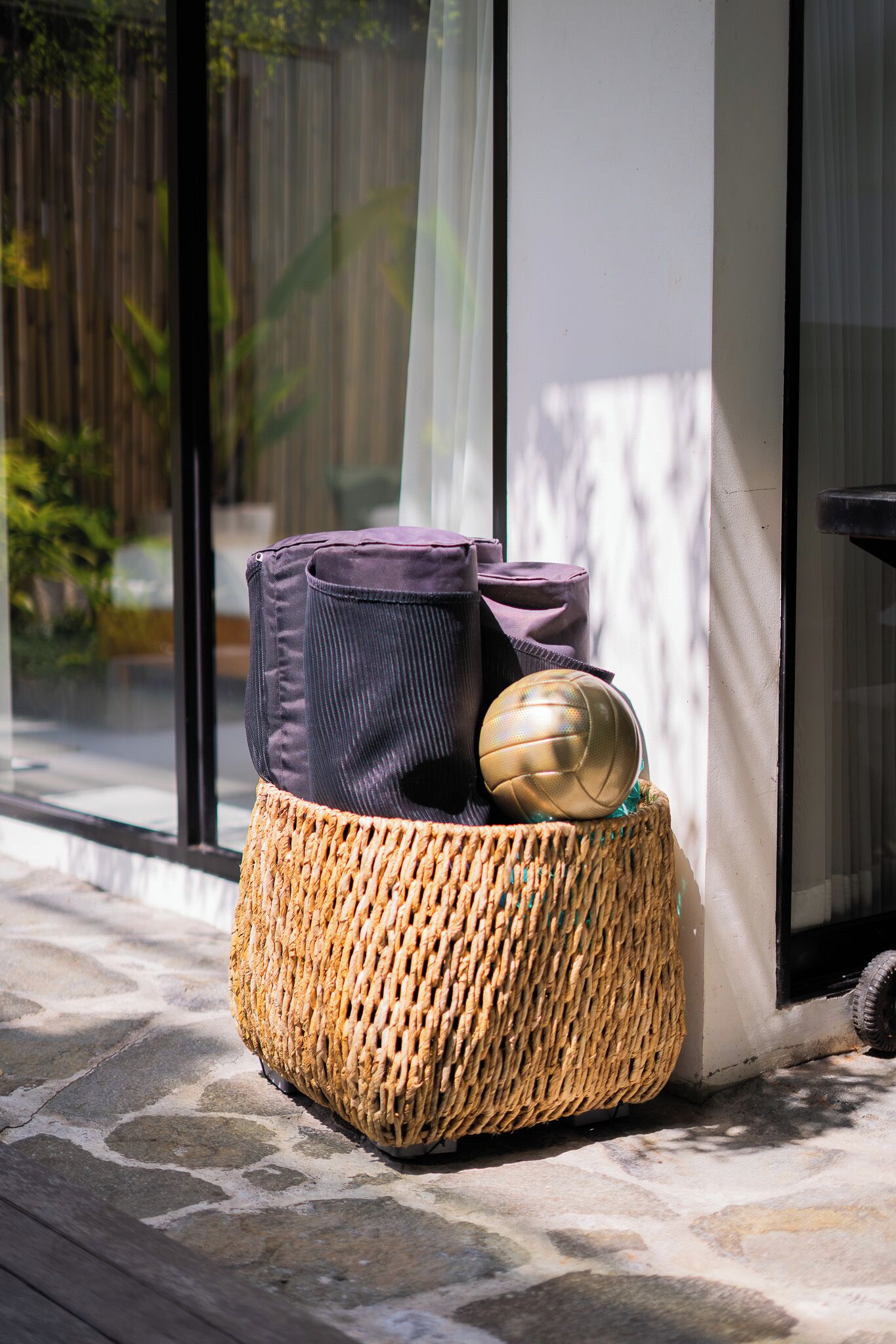 Uma cesta de vime sobre um chão de pedra perto de uma porta de vidro, contendo uma bolsa roxa, um travesseiro e uma bola esportiva.