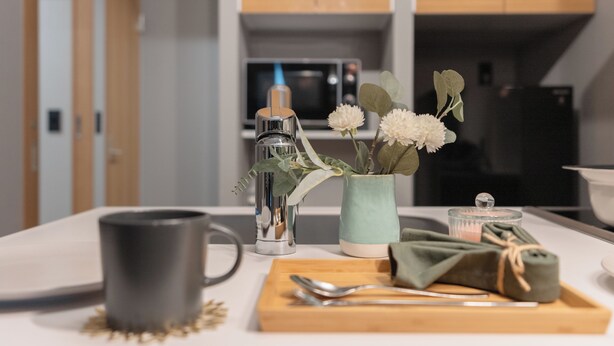 Uma bancada de cozinha com uma caneca de café, uma garrafa térmica, um vaso com flores, uma tábua de corte e um objeto embrulhado.
