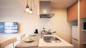 Cozinha moderna com ilha central, pia de aço inoxidável e armários de madeira. Há uma área de refeições com cadeiras e mesa, e uma TV fixada na parede.