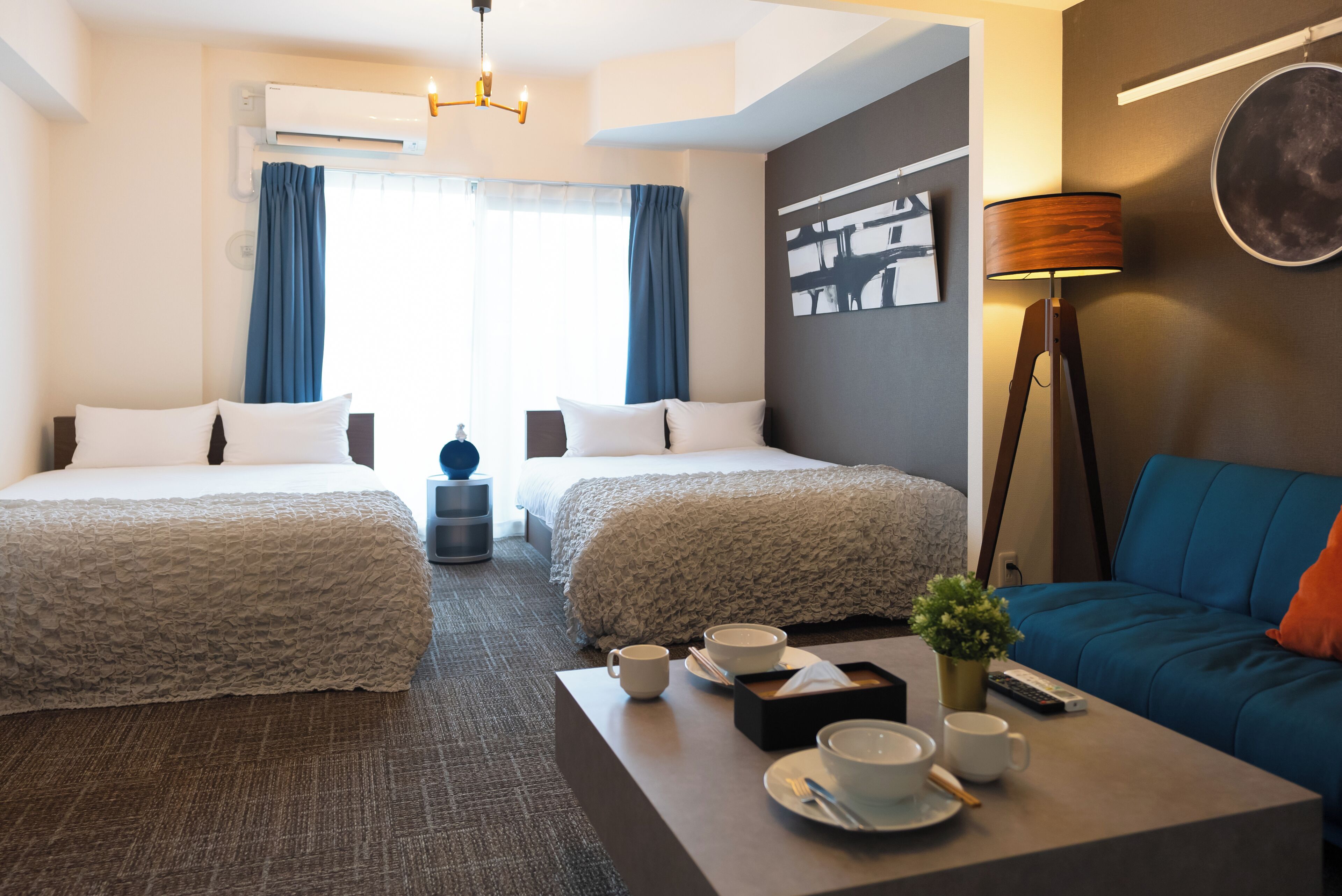 Quarto de hotel com duas camas, um sofá, uma mesa de centro com chávenas e uma planta, e um ar condicionado fixado na parede.