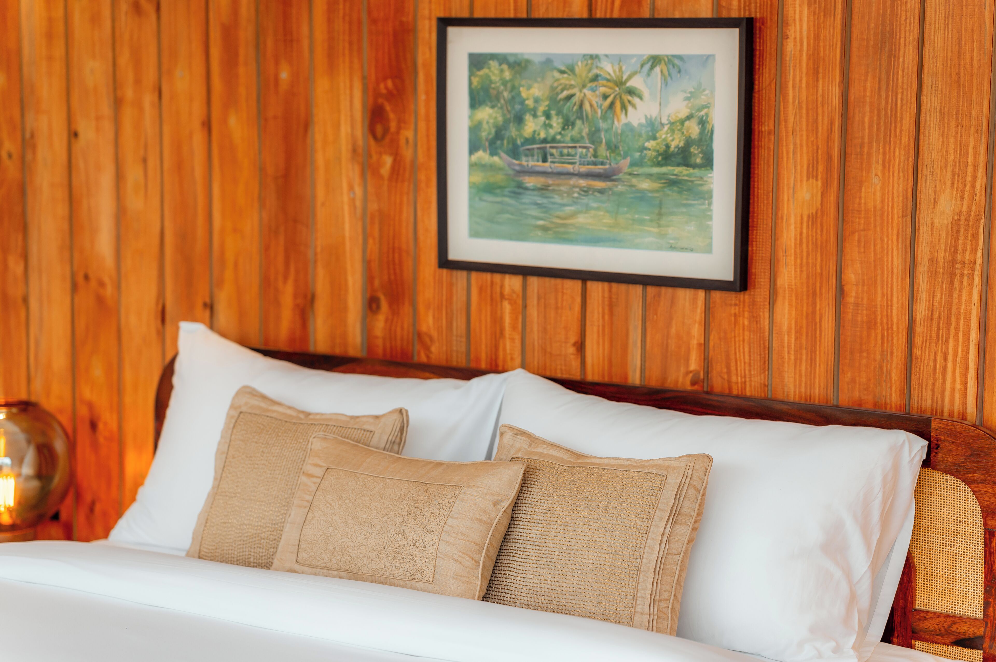 Uma cama com almofadas brancas e bege, uma cabeceira de madeira e um quadro com uma imagem de um barco na água.