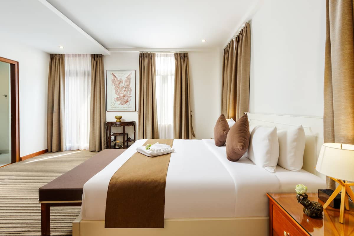 Quarto de hotel com uma cama grande, uma mesa de cabeceira com abajur, uma mesinha com objeto decorativo e um quadro na parede.