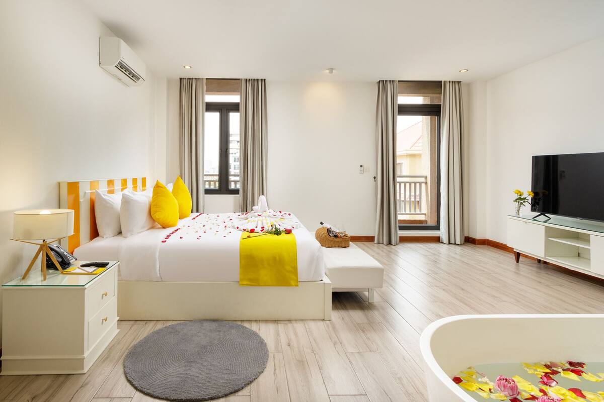 Quarto de hotel moderno com uma cama grande, uma mesinha de cabeceira com abajur, uma televisão e uma banheira com detalhes florais.