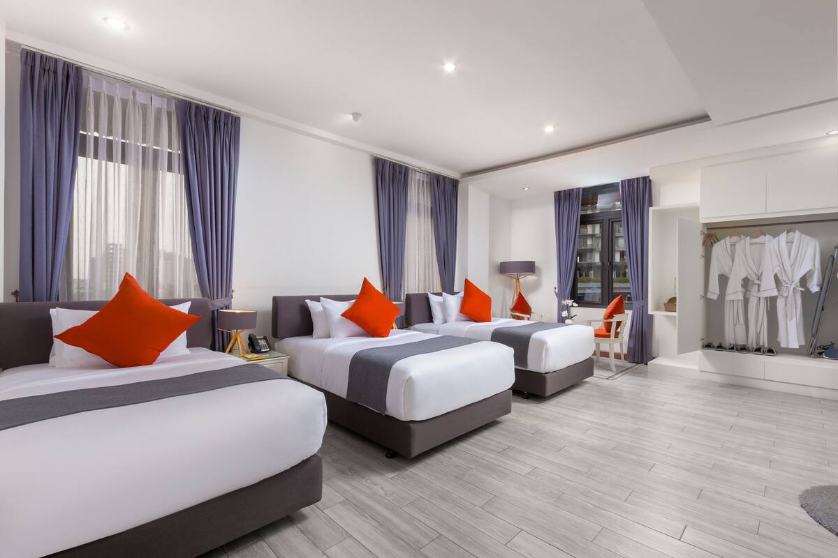 Quarto de hotel com três camas, roupão e porta-toalhas, e uma janela grande com cortinas.