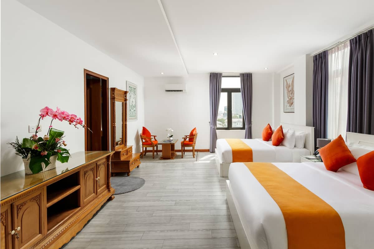Quarto de hotel com duas camas, penteadeira de madeira, escrivaninha, cadeiras e uma janela grande com cortinas.