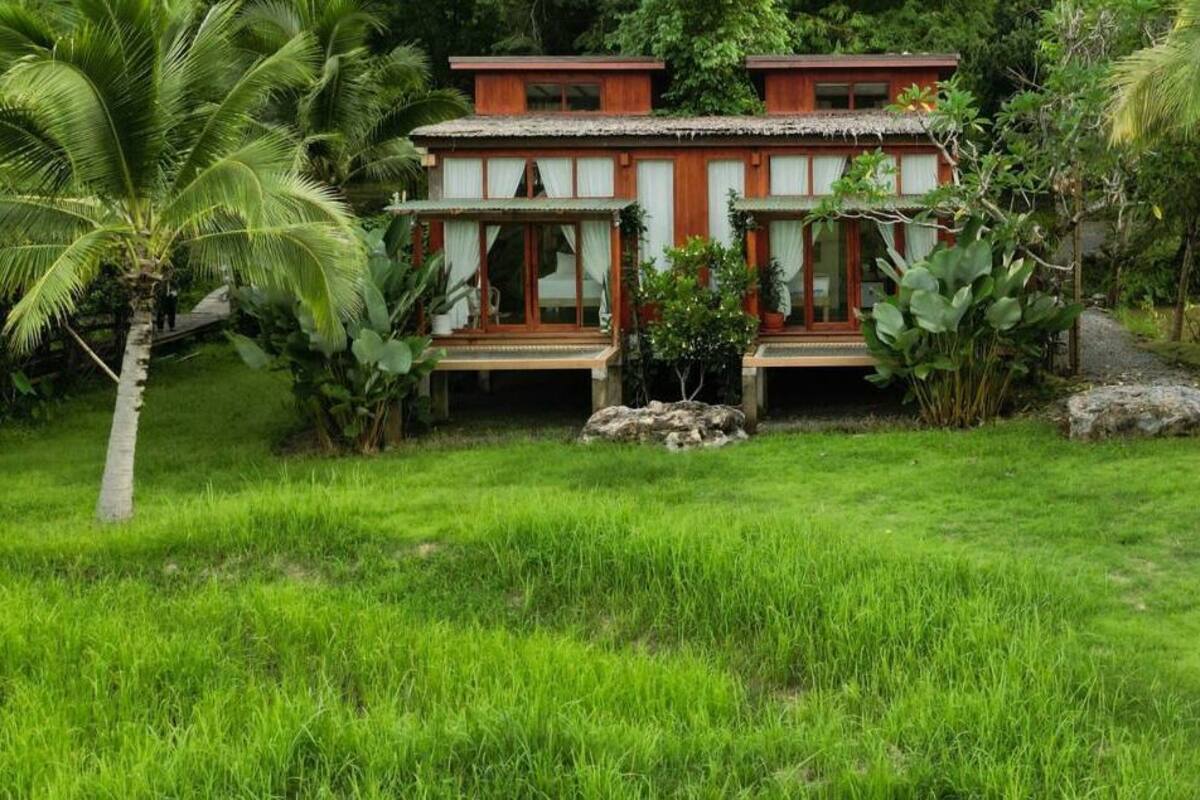 Uma casa de campo com telhado de palha, cercada por vegetação exuberante e palmeiras.
