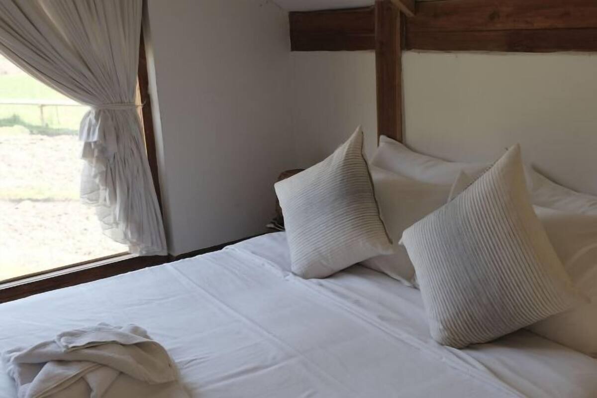 Cama com lençóis e almofadas brancas, janela com cortinas e estrutura de teto de madeira.