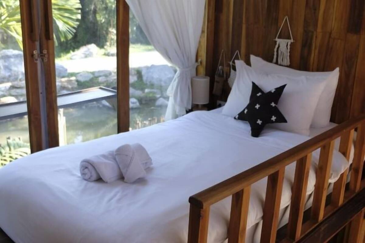 Cama com estrutura de madeira, roupa de cama branca, toalha enrolada e almofada estampada com estrelas.