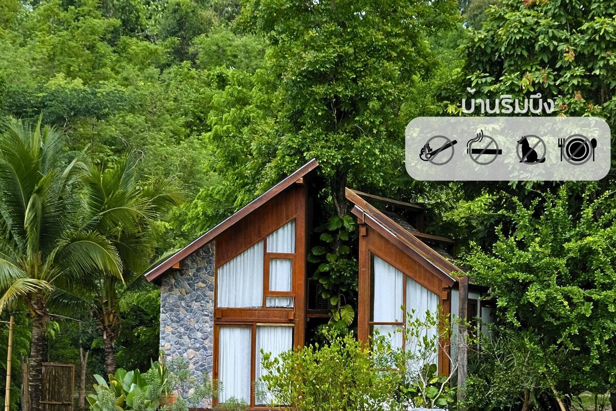 Uma casa de madeira rústica com paredes de pedra, rodeada por vegetação exuberante e uma grande rocha em primeiro plano.