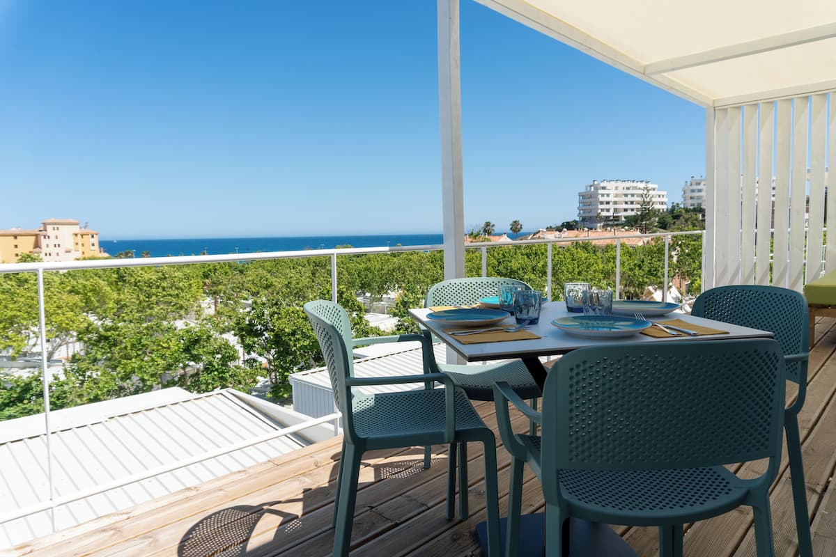Varanda com mesa posta para refeições, vista para a praia e paisagem urbana.