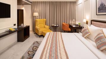 Quarto de hotel com uma cama grande, uma escrivaninha, uma poltrona amarela, uma cadeira laranja, uma mesa pequena e uma TV fixada na parede.