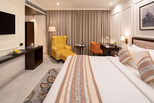 Quarto de hotel com uma cama grande, uma escrivaninha, uma poltrona amarela, uma cadeira laranja, uma mesa pequena e uma TV fixada na parede.