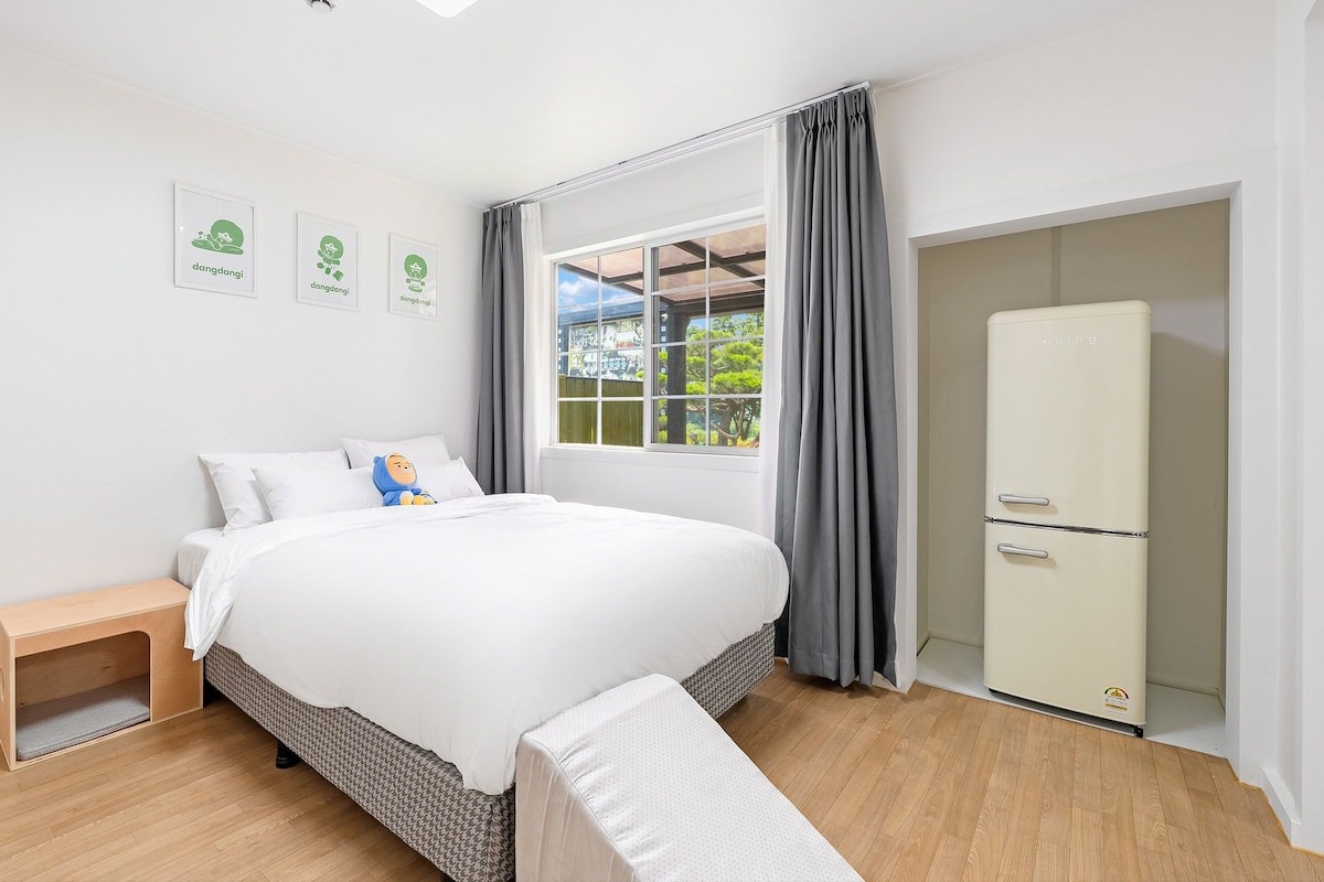Quarto com cama, mesa de cabeceira, janela com cortinas e geladeira.