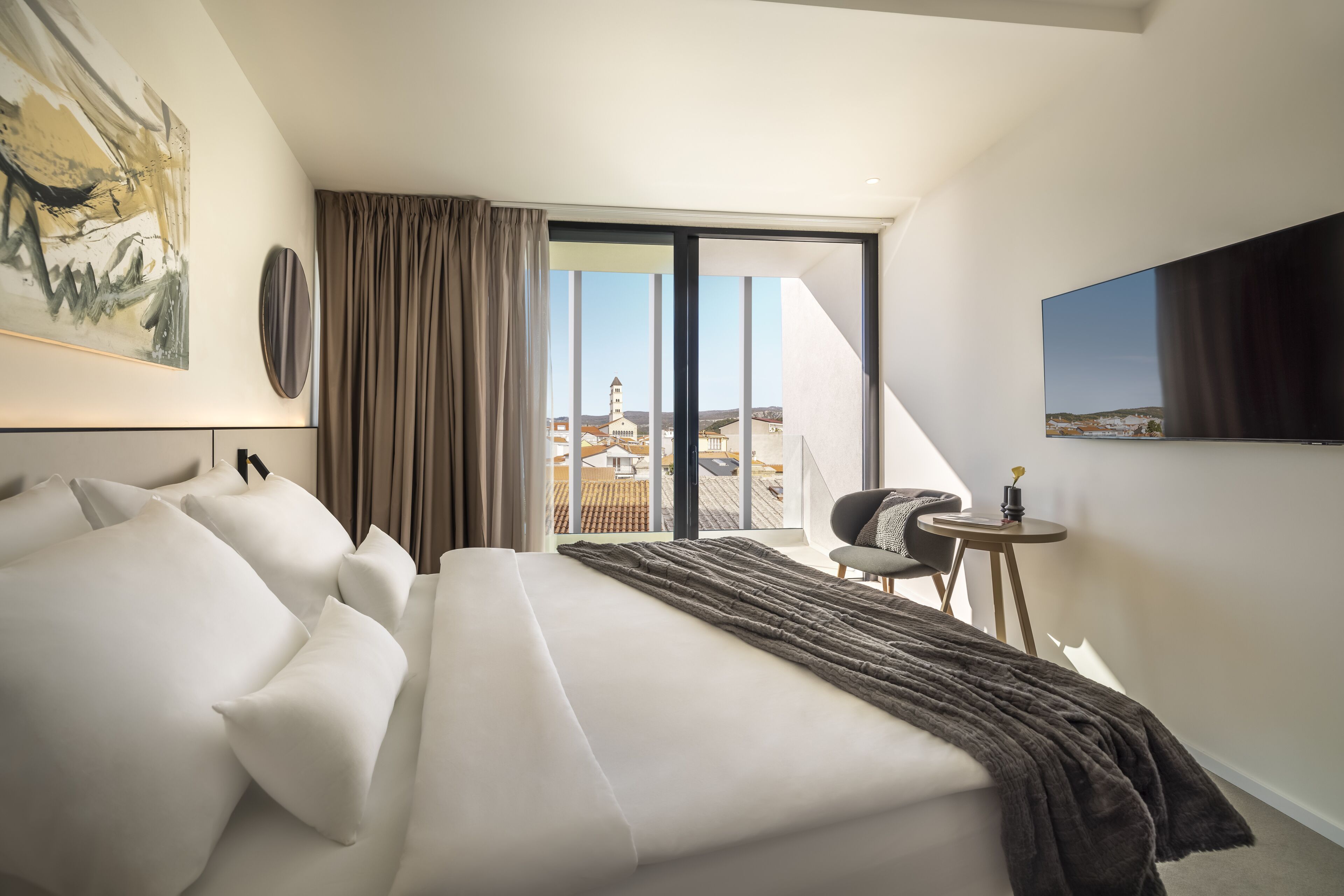 Quarto de hotel moderno com uma cama grande, vista para a cidade e uma pequena mesa com uma cadeira.